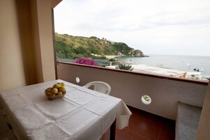 Varanda com mesa posta para refeições, com vista para a paisagem costeira, colinas e o mar.