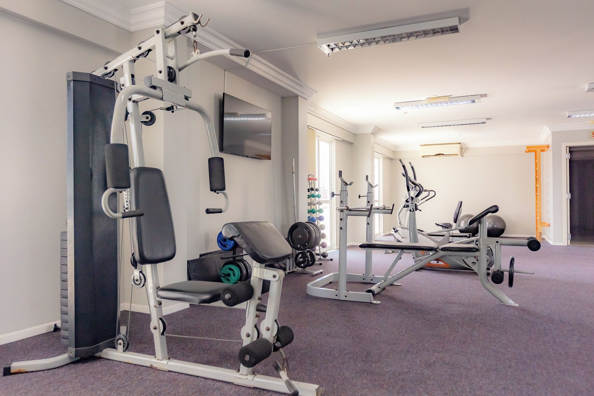 Uma academia com diversos equipamentos de exercício, uma televisão e uma porta que dá para outro cômodo.