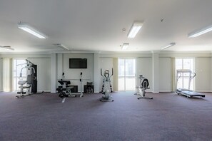 Uma academia espaçosa com diversos equipamentos de exercício, amplas janelas e uma TV de tela plana na parede.