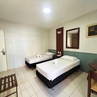Quarto de hotel com duas camas de solteiro, uma escrivaninha, uma cadeira, um espelho e uma porta.