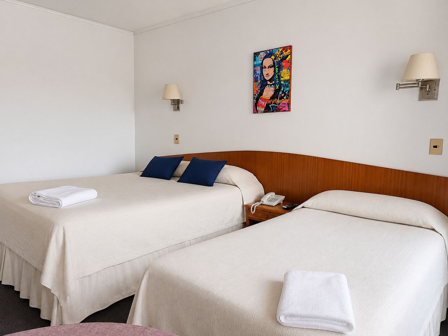 Quarto de hotel com duas camas, cabeceira de madeira, mesa de cabeceira com abajur, telefone e uma obra de arte colorida na parede.