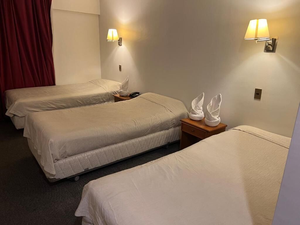 Quarto de hotel com três camas, uma cortina vermelha, uma luminária na parede e uma mesinha de cabeceira com telefone e toalhas.