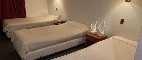 Quarto de hotel com três camas, uma cortina vermelha, uma luminária na parede e uma mesinha de cabeceira com telefone e toalhas.