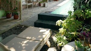 Uma área de estar coberta ao ar livre, com cadeiras e mesa de bilhar, cercada por plantas em vasos e um cercado de madeira.