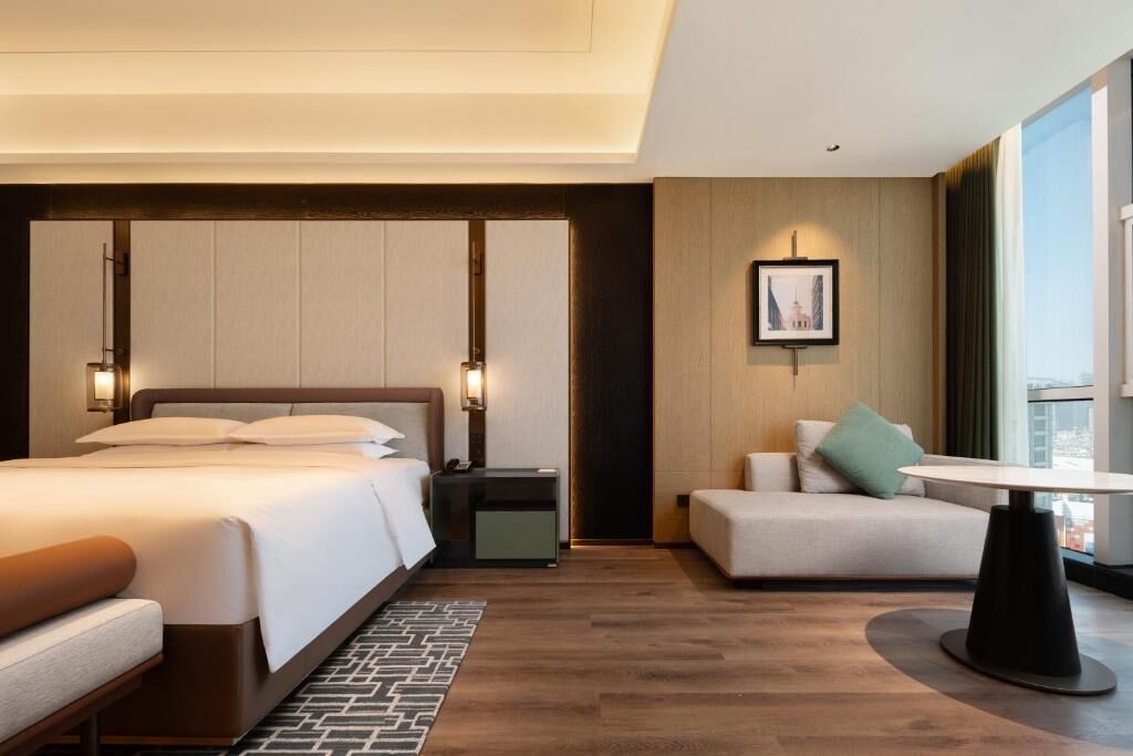 Quarto de hotel moderno com uma cama grande, área de estar com uma mesa e vista para a cidade pela janela.