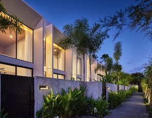 Um edifício moderno com um caminho ladeado por plantas tropicais, um portão de entrada escuro e janelas iluminadas.