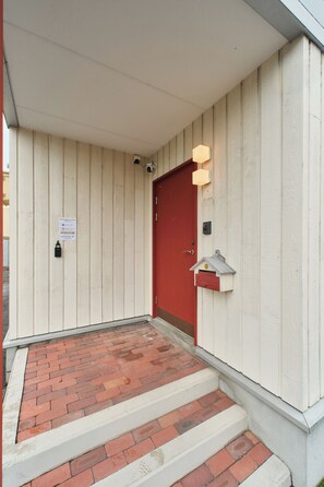 Entrada de um prédio com uma porta vermelha, uma caixa de correio e uma câmera de segurança.
