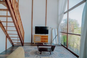 Quarto com escada de madeira, televisão de tela plana em um armário de madeira, tapete estampado e uma grande janela com vista para o verde.