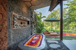 Pareda de pedra com um nicho contendo produtos de higiene, um banco de pedra com um desenho floral colorido e um caminho que leva a uma área de estar com um guarda-sol verde.