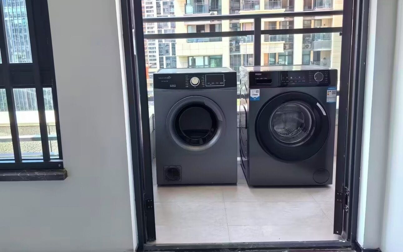 Duas máquinas de lavar roupa de abertura frontal numa lavanderia com vista para um prédio residencial através de uma janela.