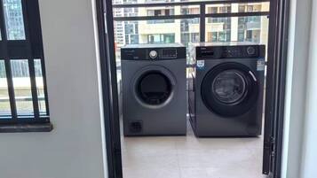 Duas máquinas de lavar roupa de abertura frontal numa lavanderia com vista para um prédio residencial através de uma janela.