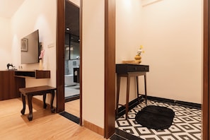 Quarto de hotel com piso de madeira, tapete com padrão preta e branca, uma mesa pequena com um vaso de flores e um banco.