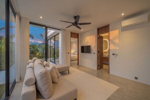 Uma sala de estar moderna com um sofá, um ventilador de teto e uma grande janela com vista para um jardim.