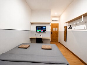 Quarto de hotel com uma cama, uma escrivaninha com monitor de computador, uma porta de madeira e um ar condicionado fixado na parede.