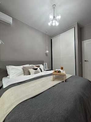 Quarto moderno com uma cama grande, mesa de cabeceira com bandeja de frutas, luminária no teto e um guarda-roupa.