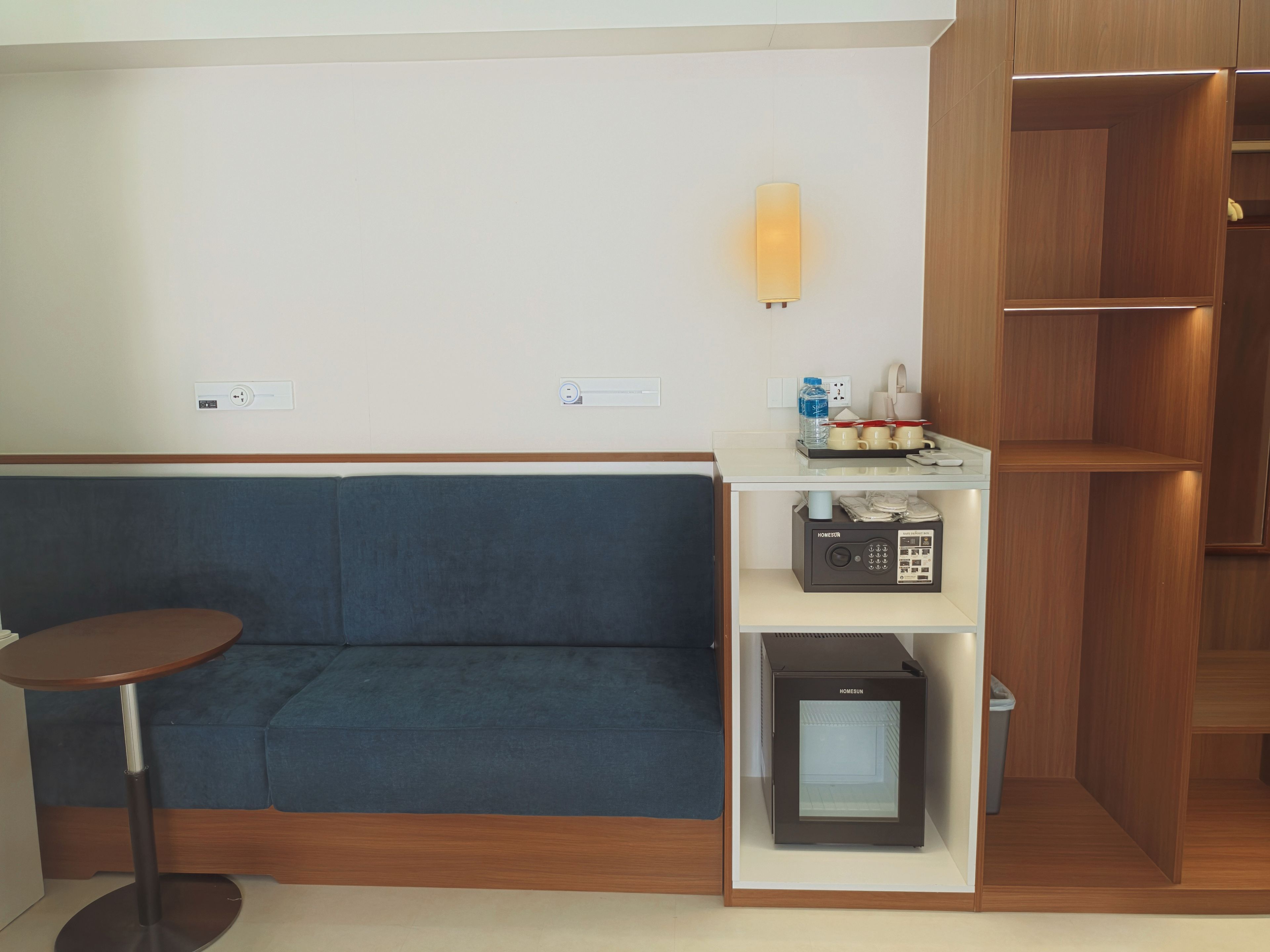 Quarto com sofá azul, mesa redonda pequena, micro-ondas e prateleira com garrafa de água e copos.