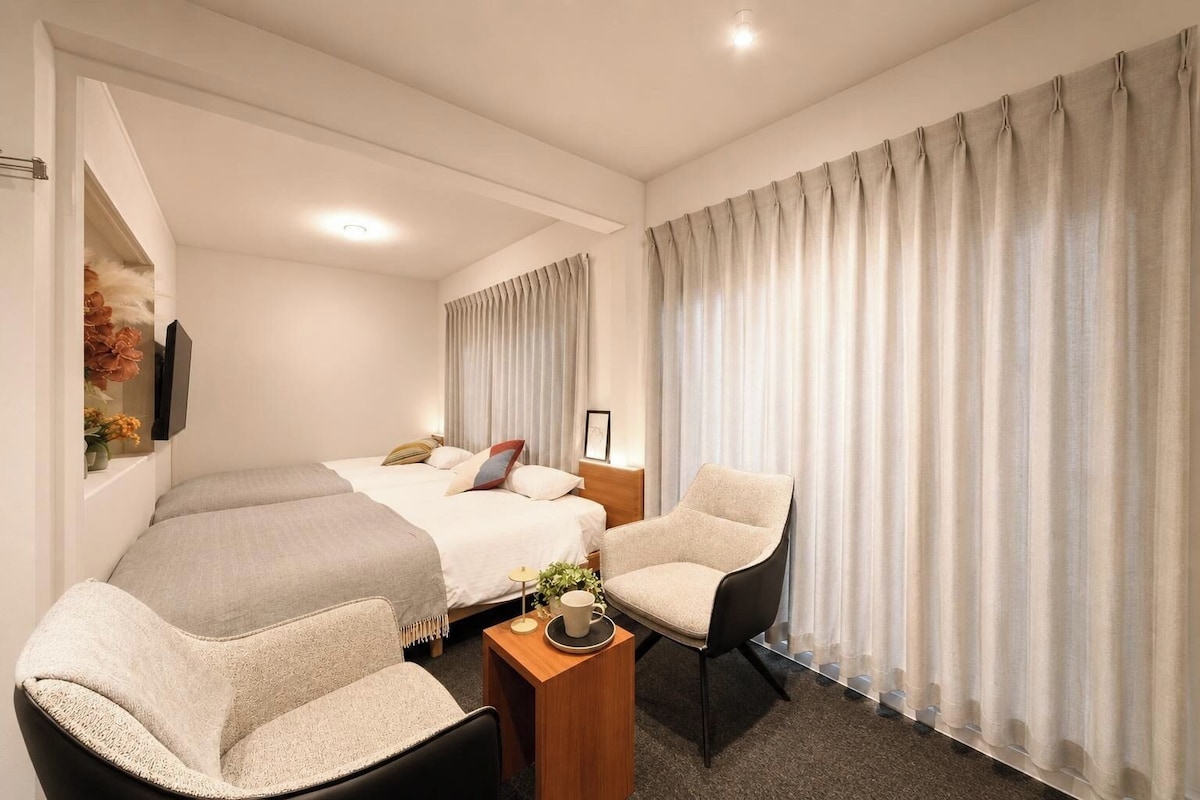 Quarto de hotel com uma cama, duas poltronas, uma mesa pequena com uma xícara e cortinas.