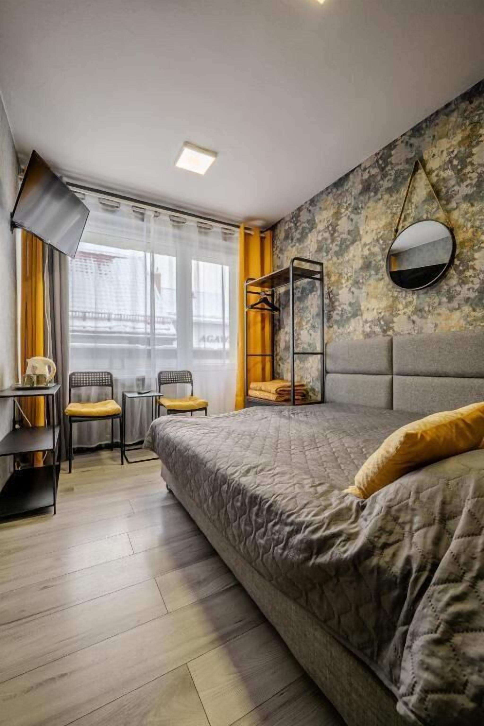 Quarto de hotel com uma cama, uma secretária com cadeira, uma mesinha e uma janela com cortinas.