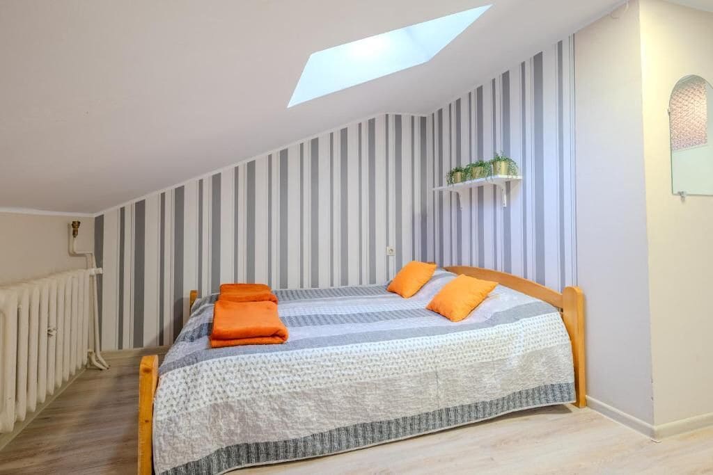 Quarto com claraboia, cama com almofadas laranja, radiador e papel de parede listrado.