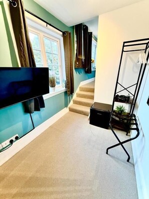 Um quarto com televisão de tela plana na parede, uma janela com cortinas, uma escada e um suporte de metal preto com objetos em cima.