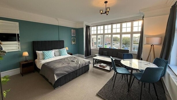 Quarto de hotel com cama, sofá, mesa de jantar com cadeiras, janela com cortinas e luminária no teto.