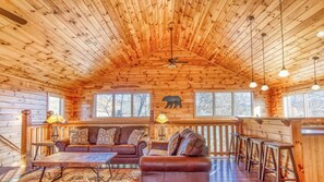 Interior de cabana de tronco com teto de madeira, um conjunto de sofás, área de jantar e uma decoração de urso na parede.