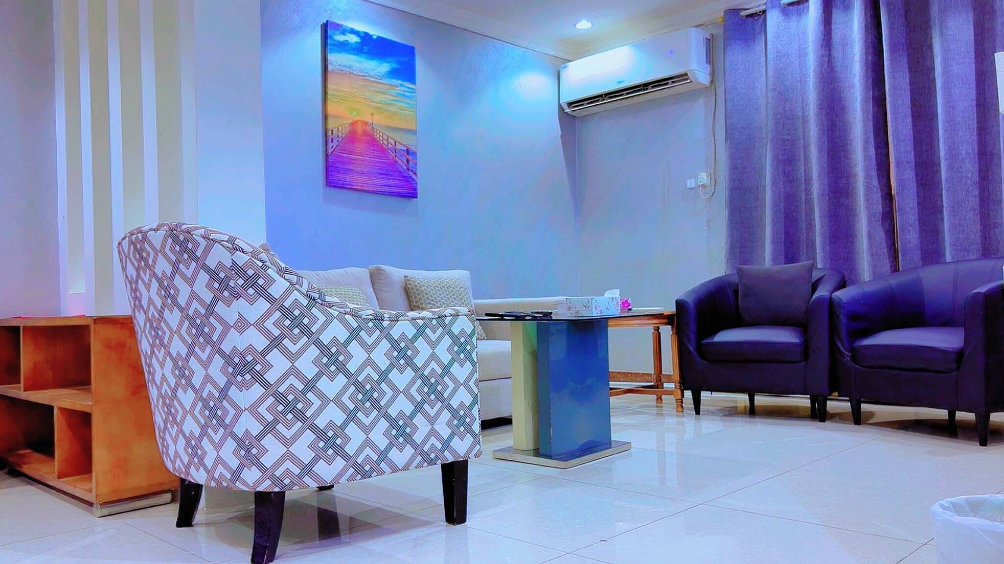 Uma sala de estar moderna com uma poltrona estampada, um sofá e duas poltronas. Há uma pintura na parede, um ar condicionado e uma janela com cortinas.