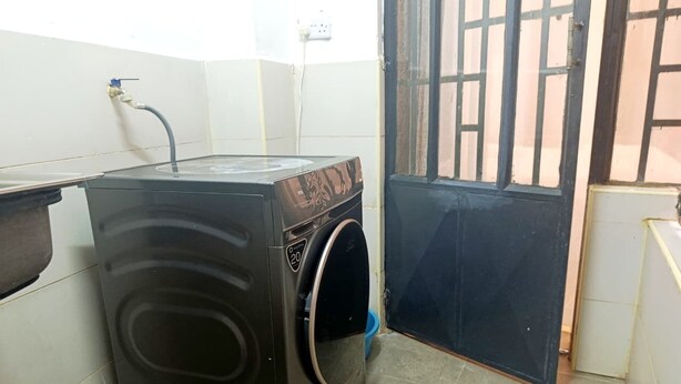 Máquina de lavar compacta num quarto pequeno com soalho em azulejos e uma porta com grelha de metal.