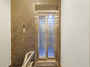 Um elevador com interior metálico e porta de vidro, localizado em um corredor com parede texturizada.