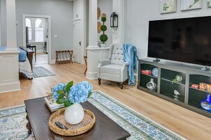 Uma sala de estar com televisão, uma poltrona, uma mesa de centro com um vaso e flores, e uma estante de livros.