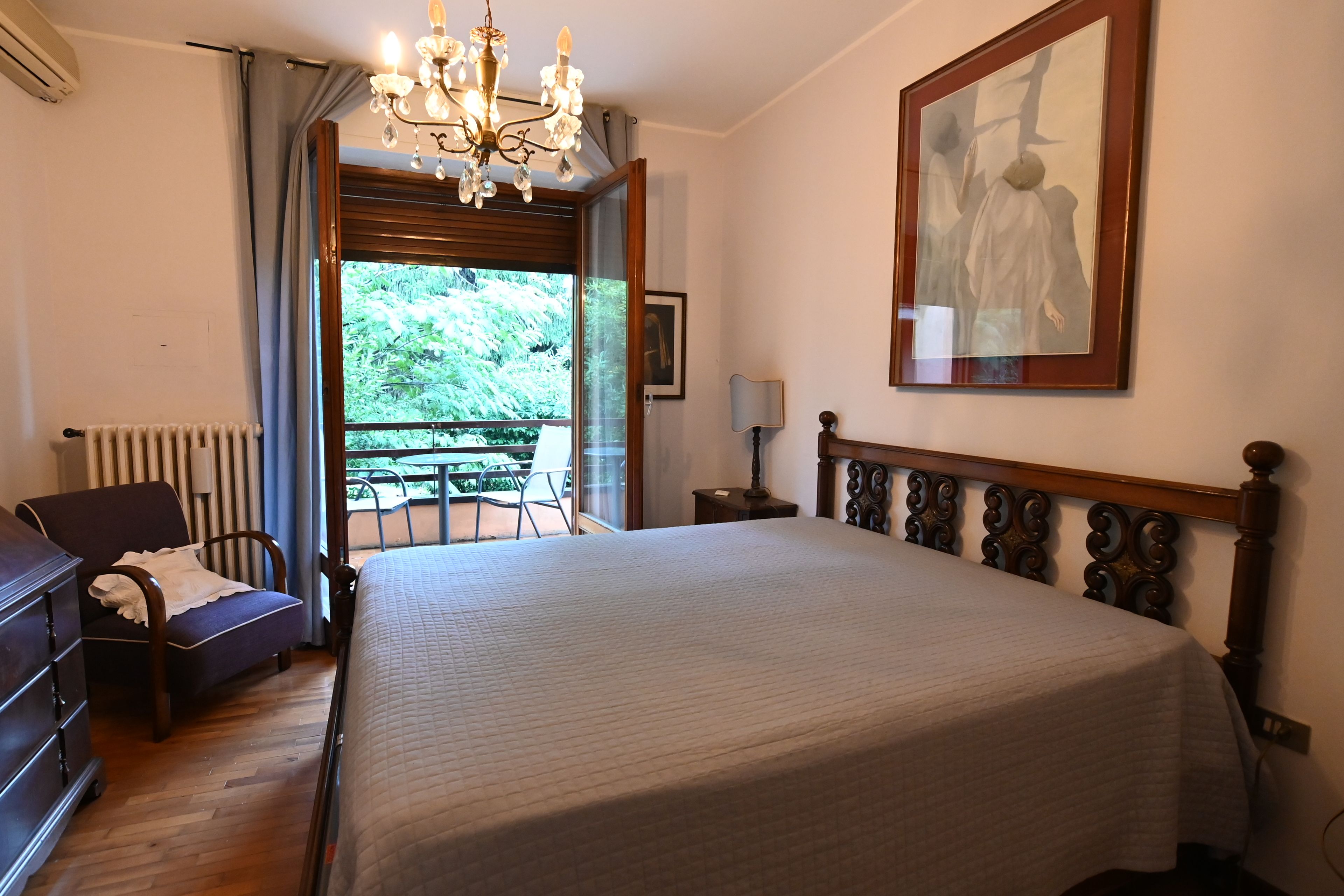 Quarto com cama grande, cabeceira de madeira, lustre, radiador, cadeira e vista para o verde lá fora.