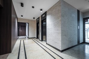 Um corredor moderno com piso de mármore, portas de madeira escura e um elevador de vidro.