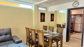 Uma sala de jantar com uma mesa posta para quatro pessoas, cadeiras de madeira, um sofá cinza e um relógio na parede.