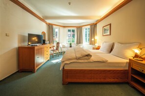 Quarto de hotel com cama, escrivaninha com televisão, cadeira, mesa de cabeceira com abajur e janela com cortinas.