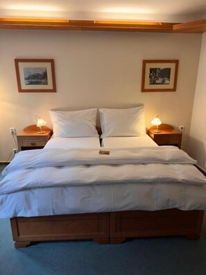 Uma cama de casal com estrutura de madeira, duas mesas de cabeceira com abajures e duas quadros nas paredes.
