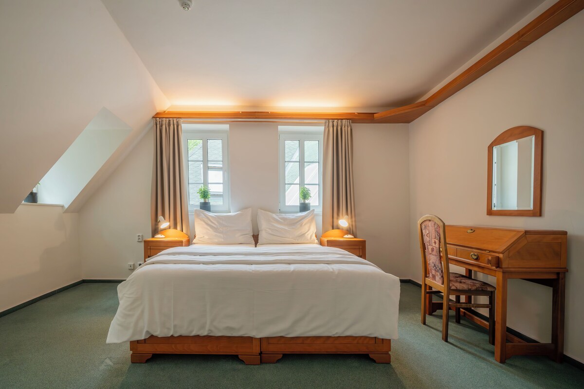 Quarto com cama, duas luminárias de cabeceira, escrivaninha com cadeira, espelho e janela com cortinas.