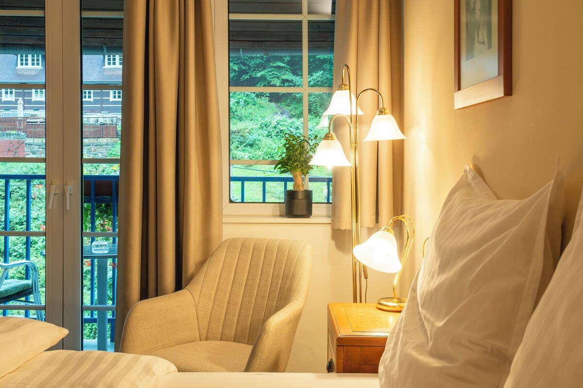 Quarto de hotel com cama, cadeira, mesa de cabeceira com luminárias, janela com cortinas e vista para o verde lá fora.