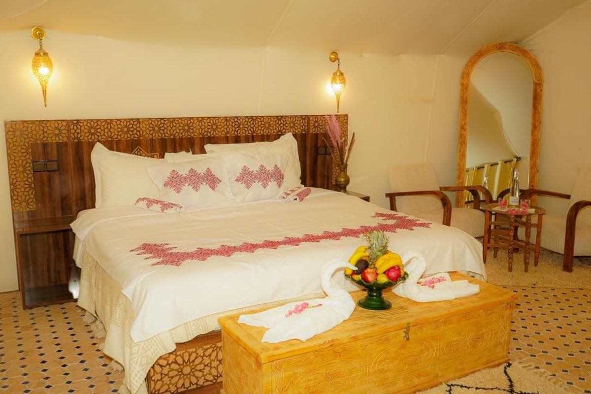 Quarto de hotel com cama grande, duas cadeiras, um baú de madeira e um espelho na parede.