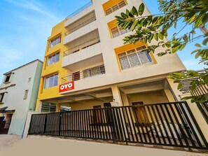 Um edifício de vários andares com fachada amarela e branca, cercado por uma grade de metal preta, e uma placa com a inscrição 'OYO ROOMS', indicando um hotel ou estabelecimento de hospedagem.