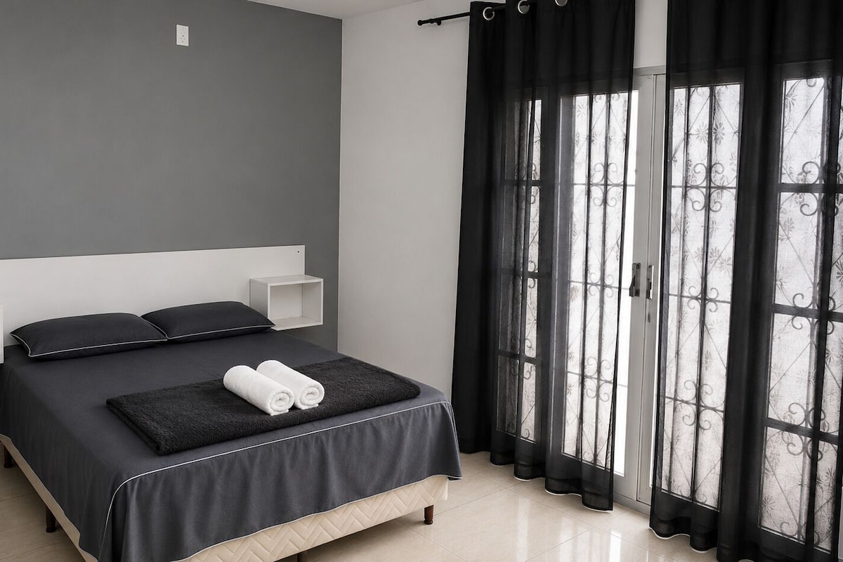 Quarto moderno com cama, cabeceira, janela com cortinas e prateleira fixada na parede.