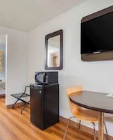 Quarto compacto com televisão de tela plana na parede, uma pequena geladeira preta, uma mesa de jantar de madeira com uma única cadeira e uma cadeira dobrável de metal.