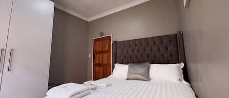 Cama bem arrumada com almofadas e toalha dobrada, guarda-roupa de madeira e porta fechada em quarto de hotel.