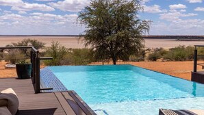 Uma piscina retangular com deck de madeira, rodeada por vegetação do deserto e um céu claro.