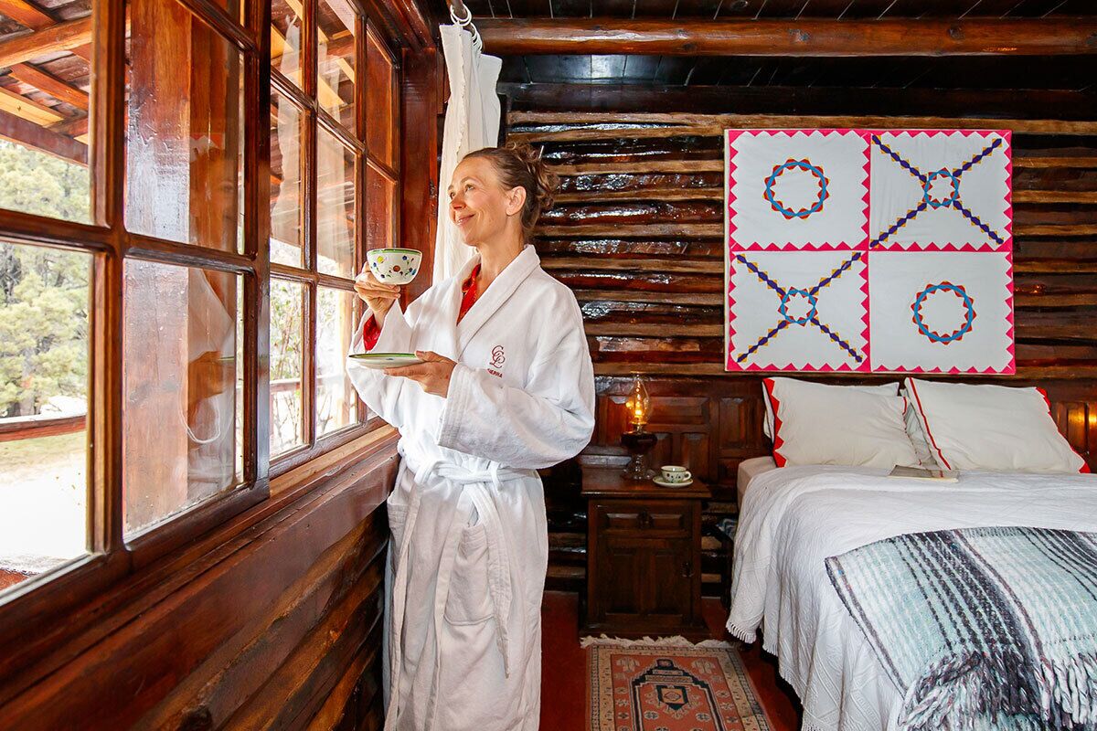 Interior de uma cabana de madeira com uma cama, um colcha e uma pessoa segurando uma xícara.