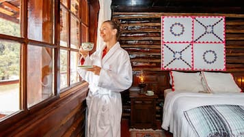Interior de uma cabana de madeira com uma cama, um colcha e uma pessoa segurando uma xícara.