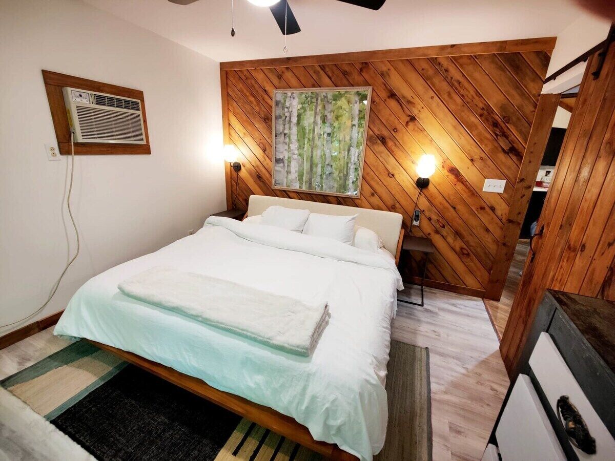 Quarto com cama, luminárias de cabeceira, painel de parede em madeira e um quadro emoldurado.