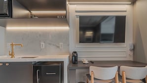 Uma cozinha compacta equipada com micro-ondas embutido, máquina de lavar louça em aço inoxidável e máquina de café. Há uma pequena mesa com duas cadeiras e uma janela acima.