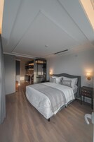 Quarto moderno com uma cama grande, mesas de cabeceira, um guarda-roupa e um banheiro visível através de uma porta aberta.