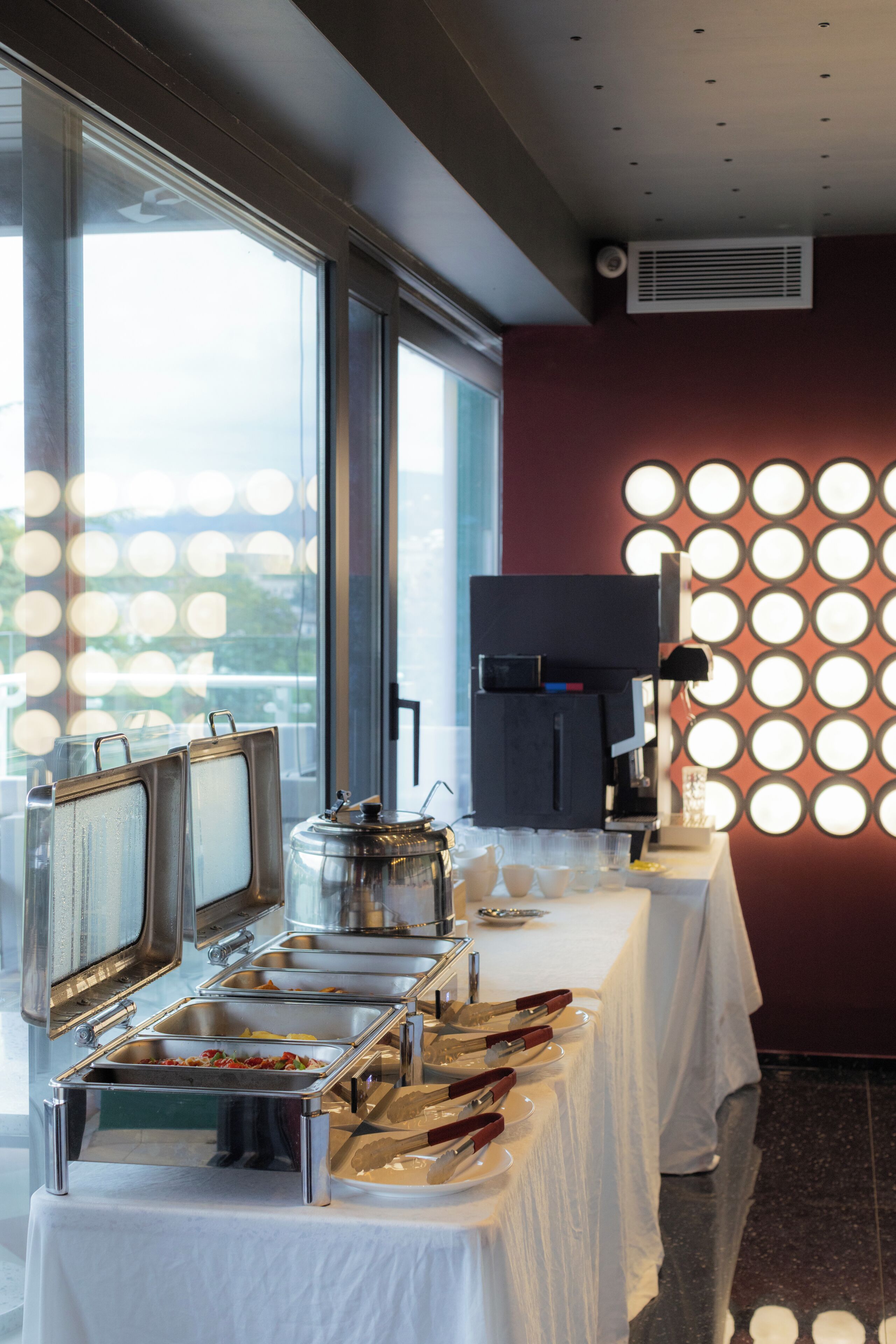 Disposição de buffet com várias estações de serviço, uma máquina de café e uma parede com luminárias circulares.