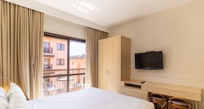 Quarto de hotel com cama, televisão, guarda-roupa de madeira, escrivaninha e varanda com cadeiras.